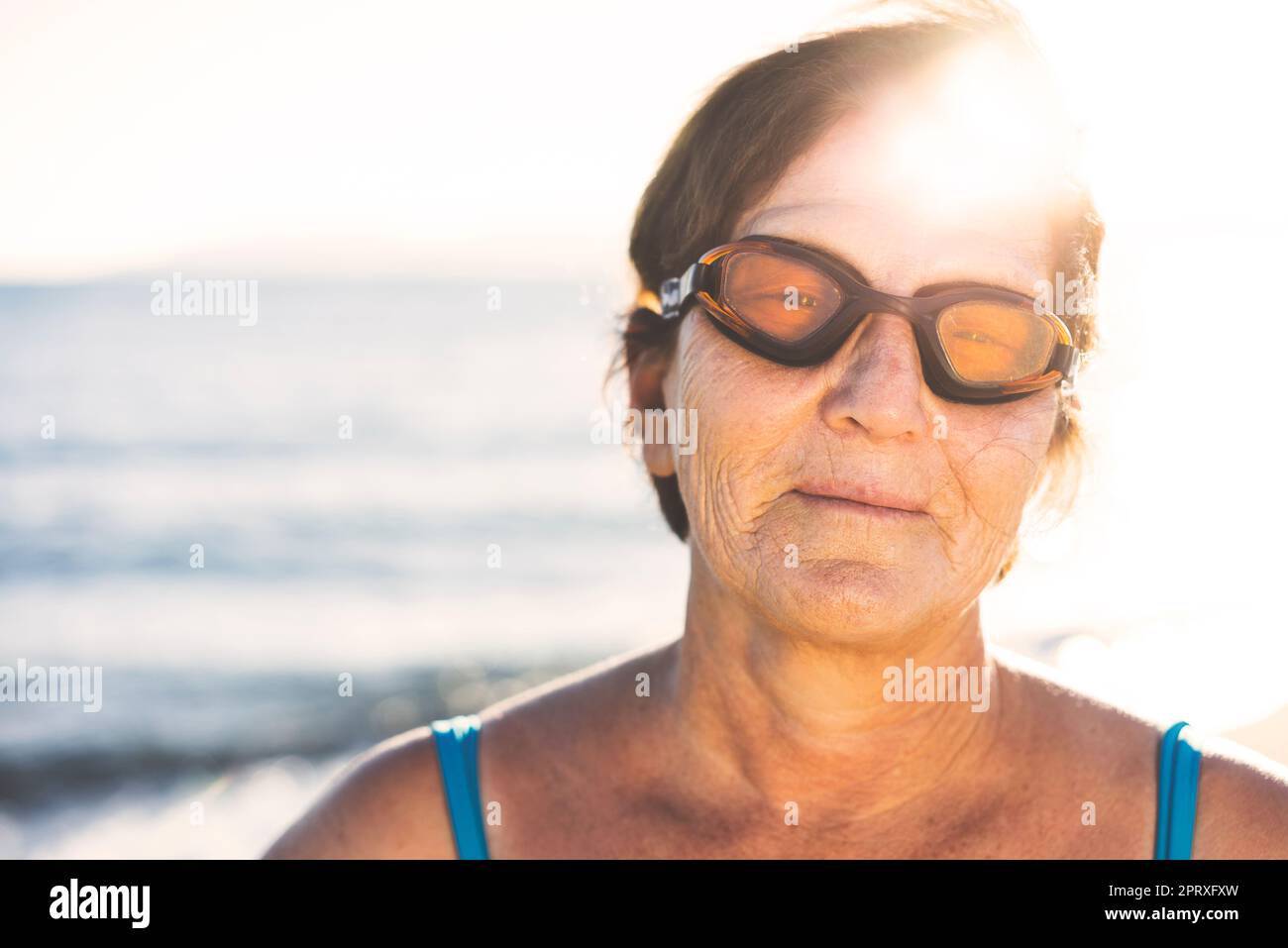 Senior woman at the beach, standing by the sea. Wearing a blue swimsuit ...