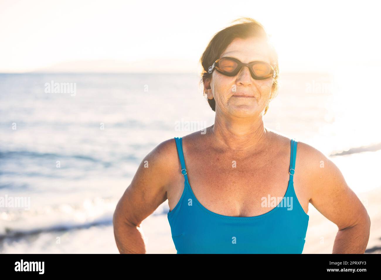 Senior woman at the beach, standing by the sea. Wearing a blue swimsuit ...