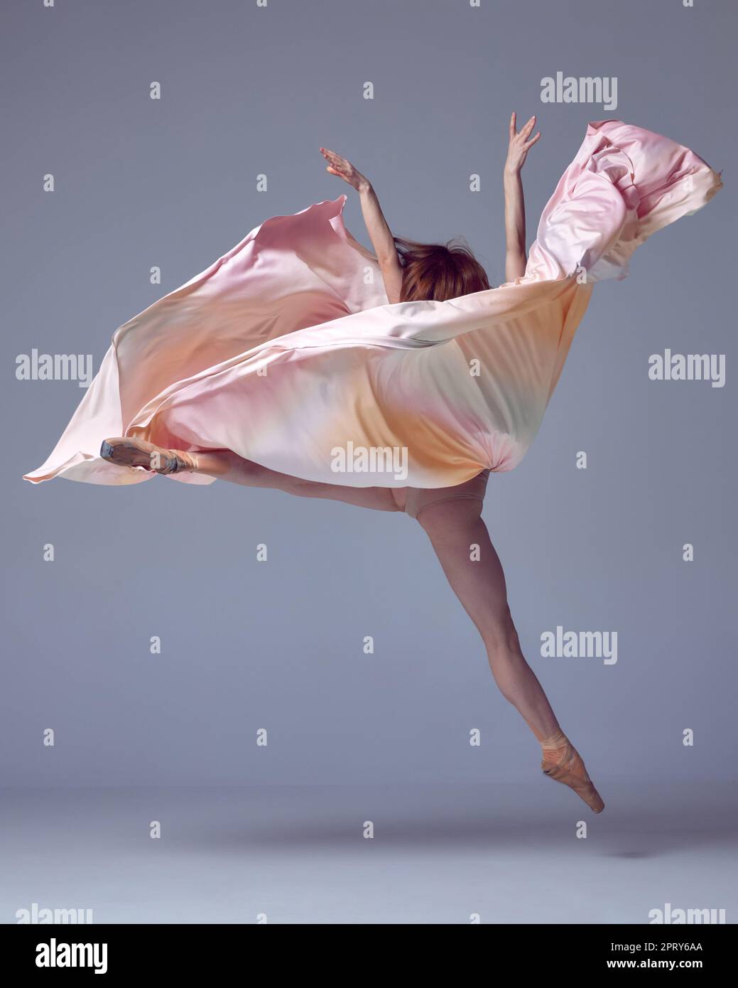 One charming professional ballerina wearing silk dress jumping up over grey studio background ...