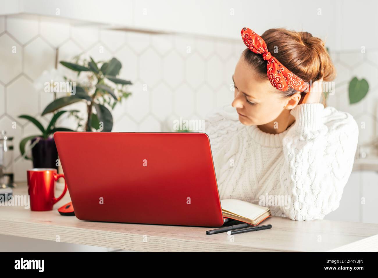 Young woman sits with red laptop at home in kitchen in red bandage and ...