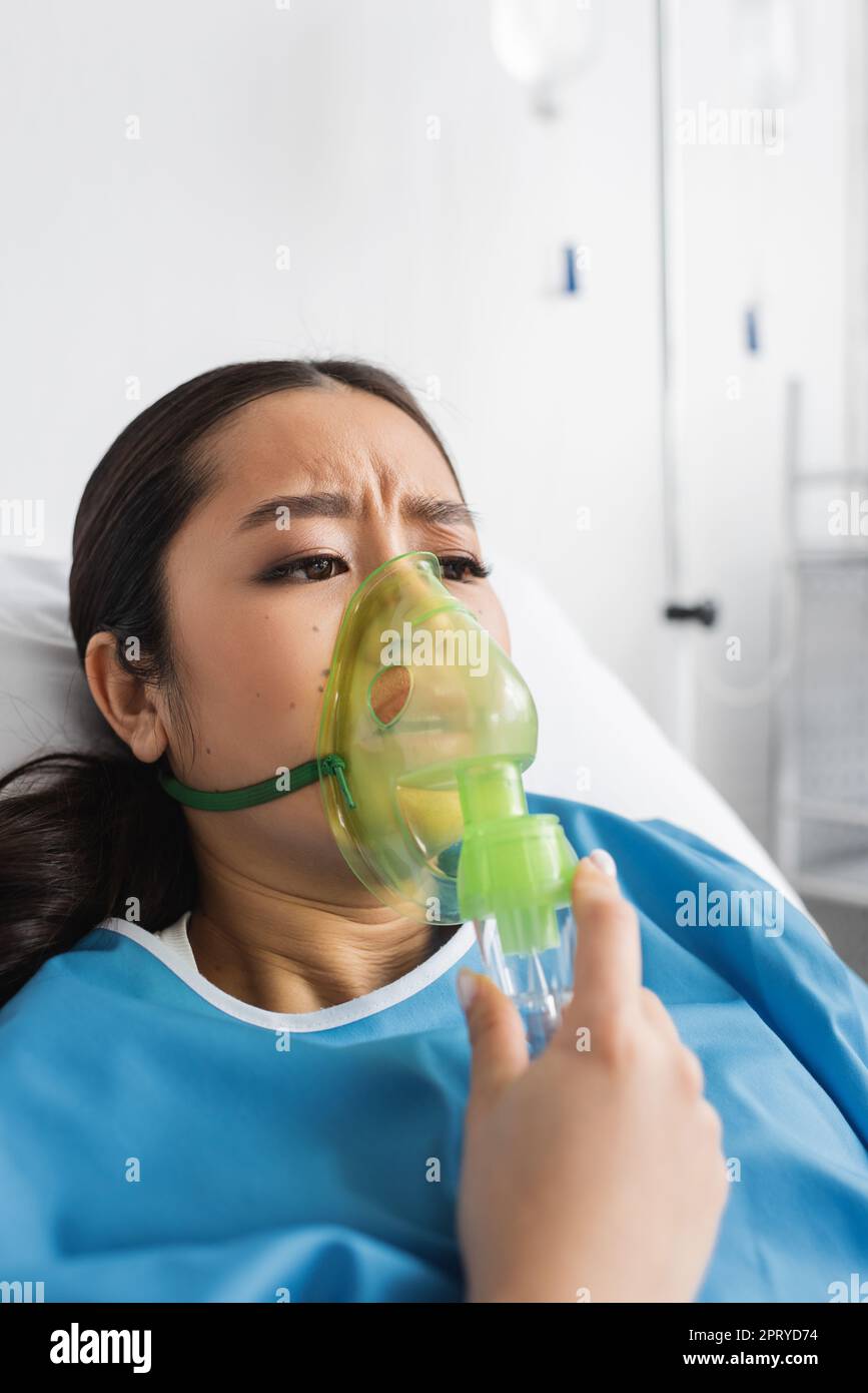 worried asian woman breathing in oxygen mask while lying on bed in ...