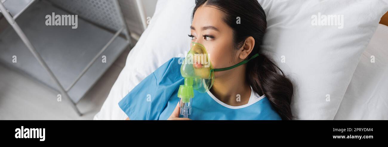 top view of depressed asian woman in oxygen mask looking away on bed in hospital ward, banner ...