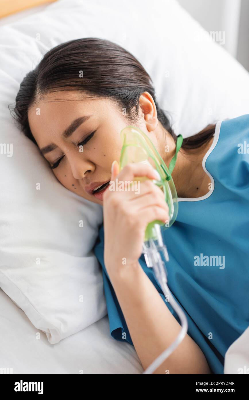 sick asian woman holding oxygen mask while lying on hospital bed with ...