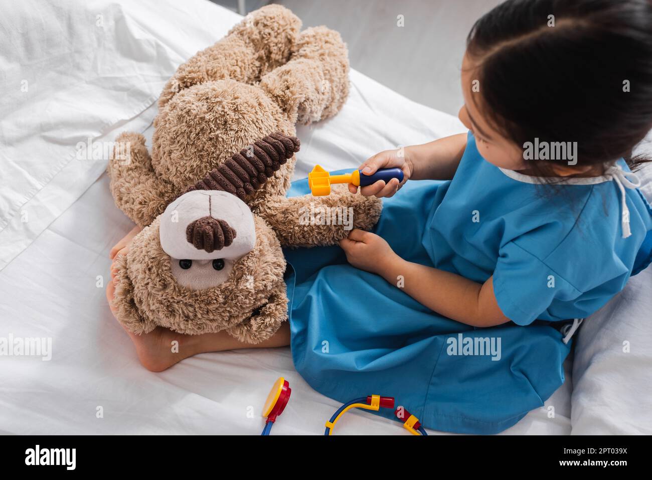 top view of asian kid in hospital gown holding toy reflex hammer near ...