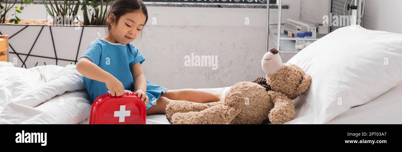asian child opening toy first aid kid near teddy bear on hospital bed ...