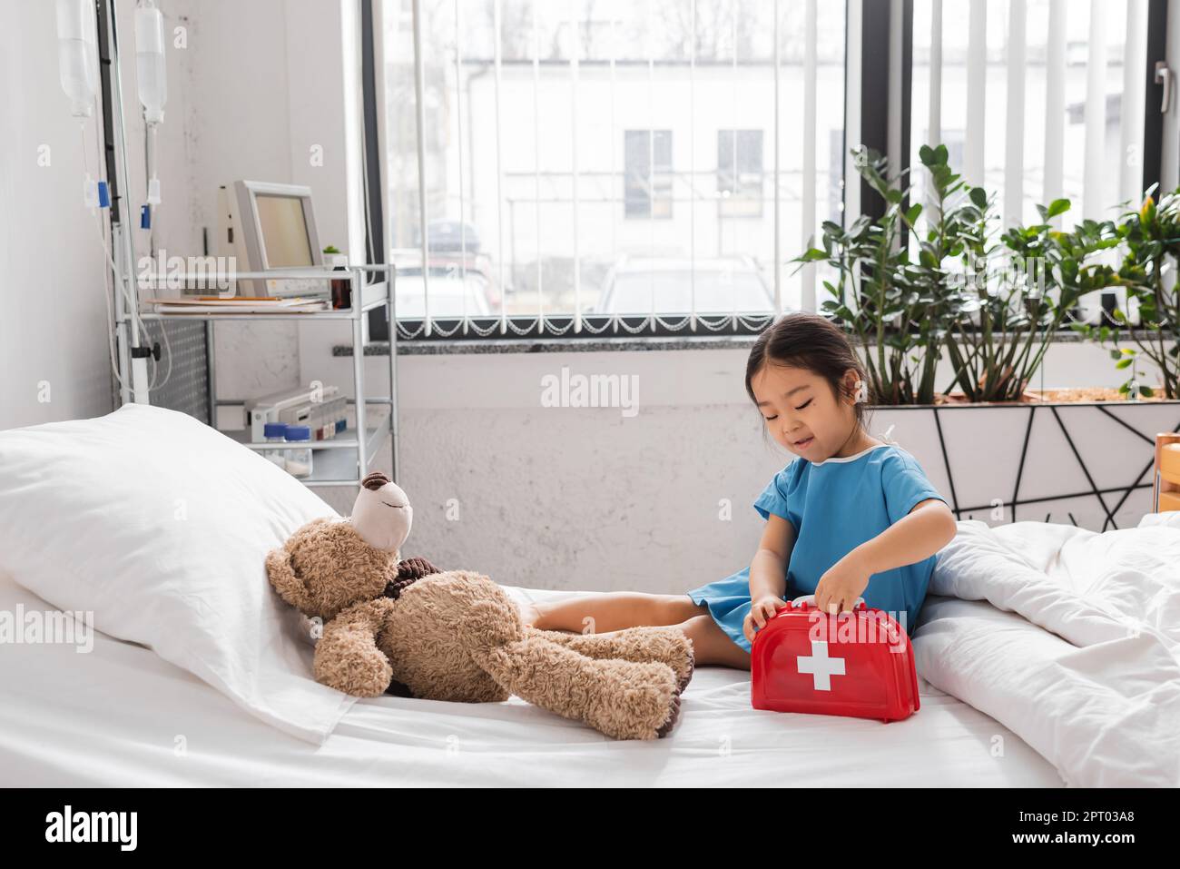 asian girl opening toy first aid kit near teddy bear on bed in modern ...