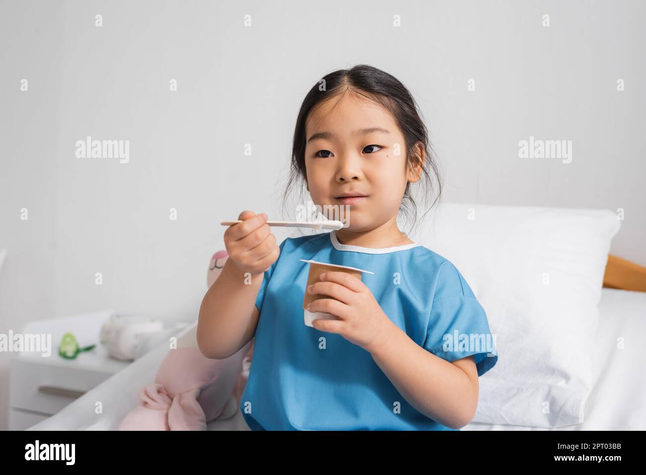 positive asian child in hospital gown eating delicious yogurt in clinic ...
