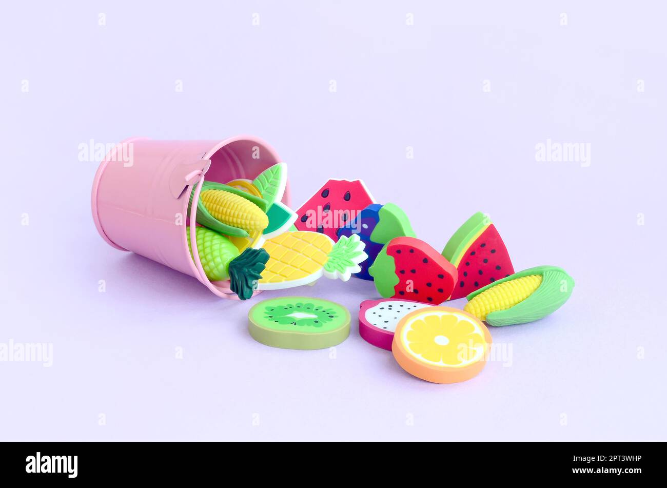 Various fresh fruits are poured from a small pink pail on violet ...