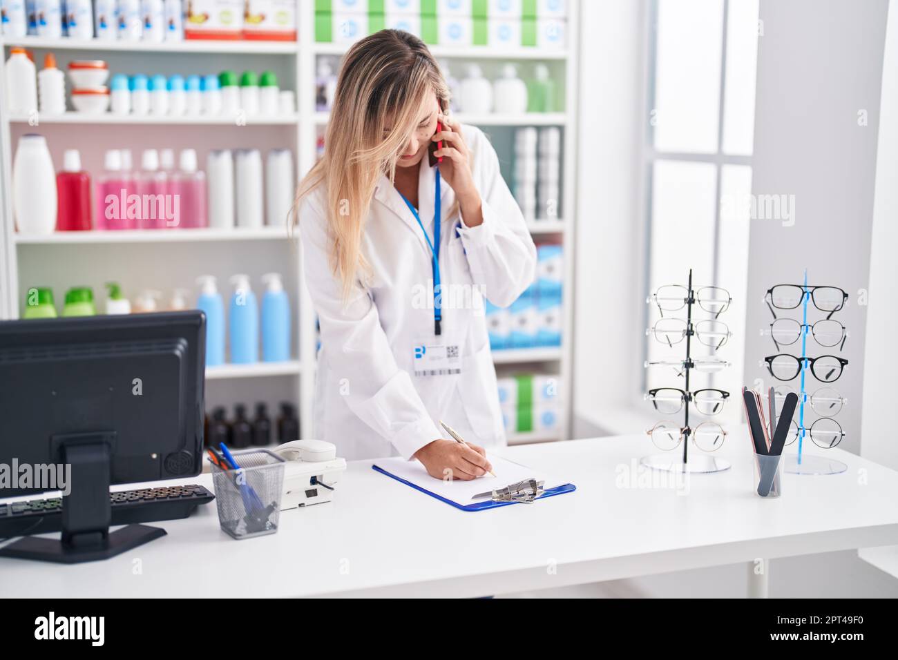 Young blonde woman pharmacist talking on smartphone writing on document ...