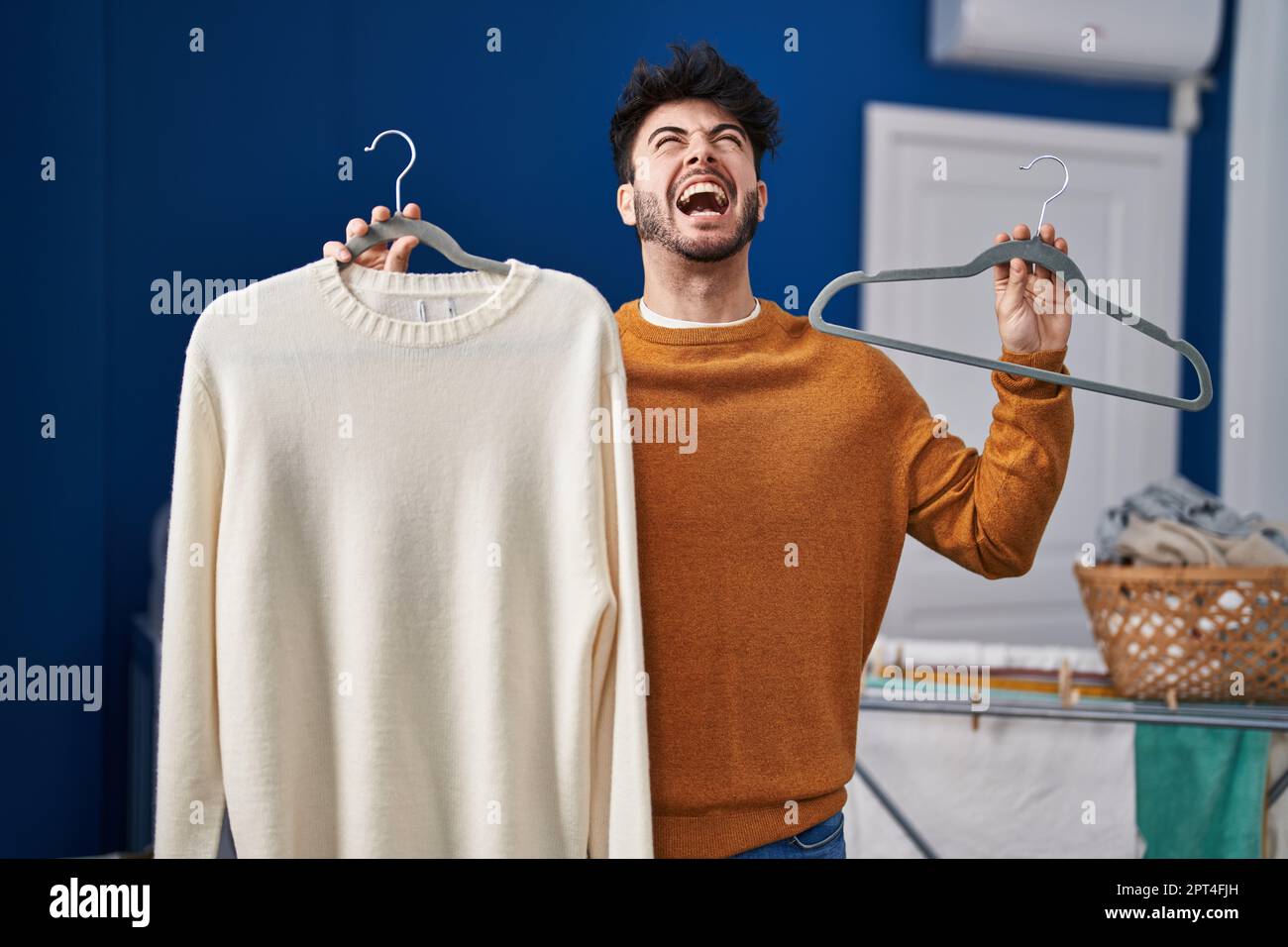 Hispanic man with beard holding sweater on hanger at laundry room angry ...