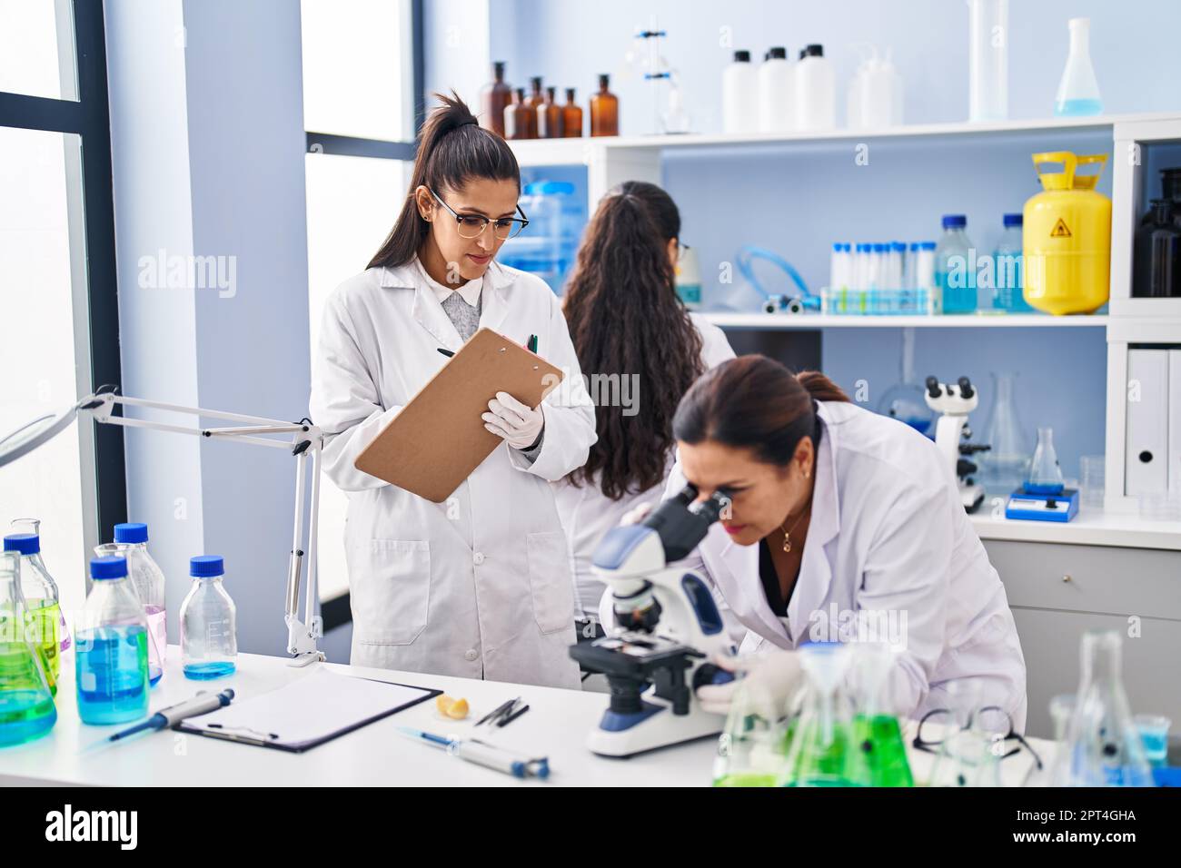 Three woman scientists using microscope write on checklist at ...