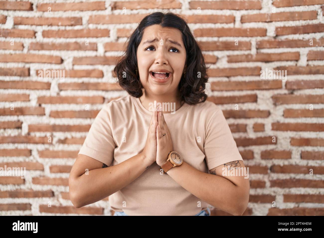 Young hispanic woman standing over bricks wall begging and praying with ...