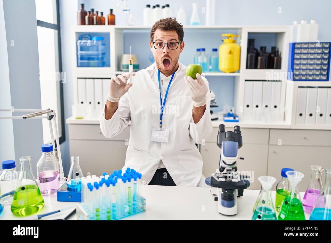 Young hispanic man working at scientist laboratory holding apple afraid ...