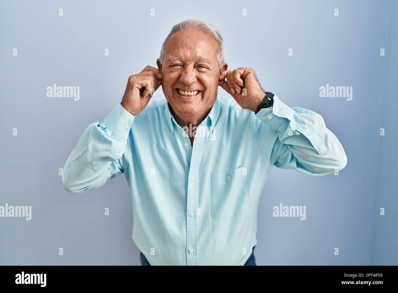 Senior man with grey hair standing over blue background covering ears ...