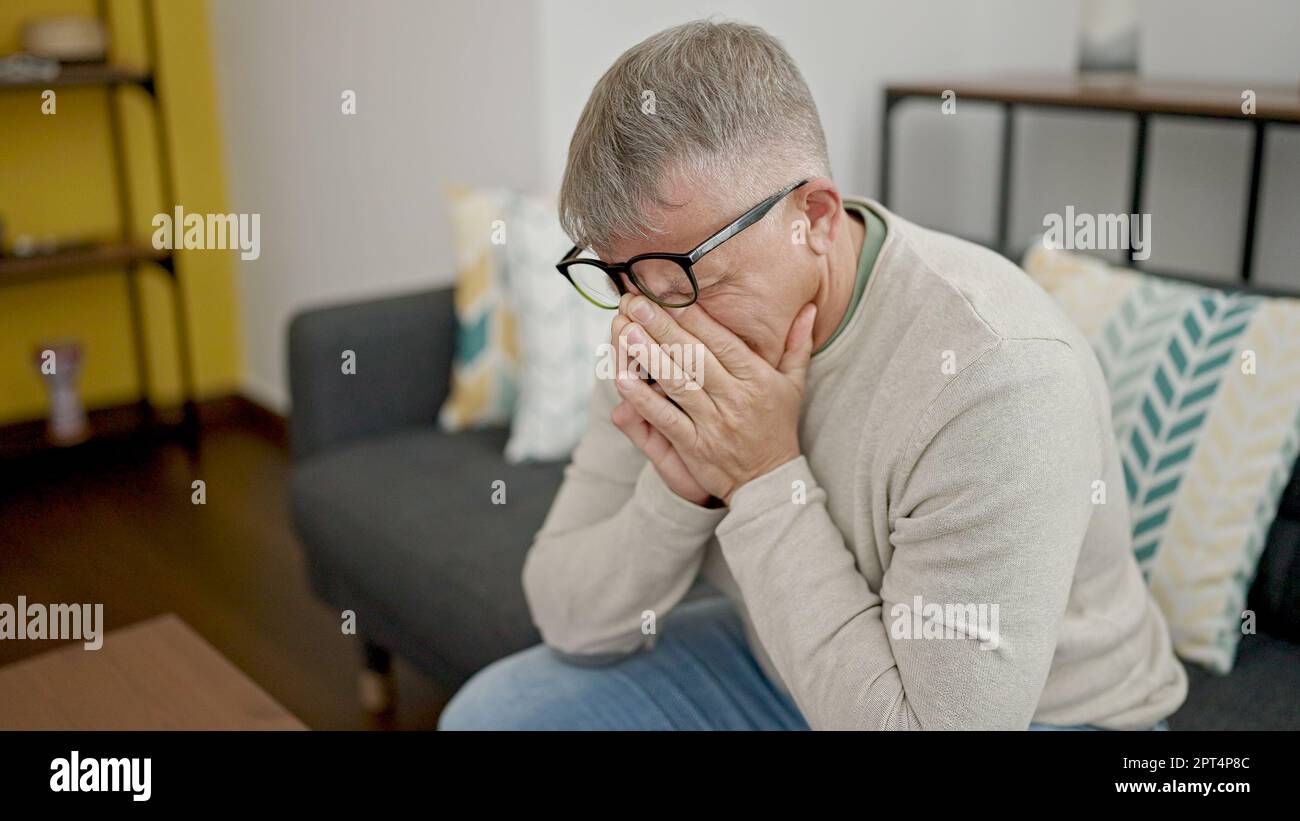 Middle age grey-haired man stressed sitting on sofa crying at home ...