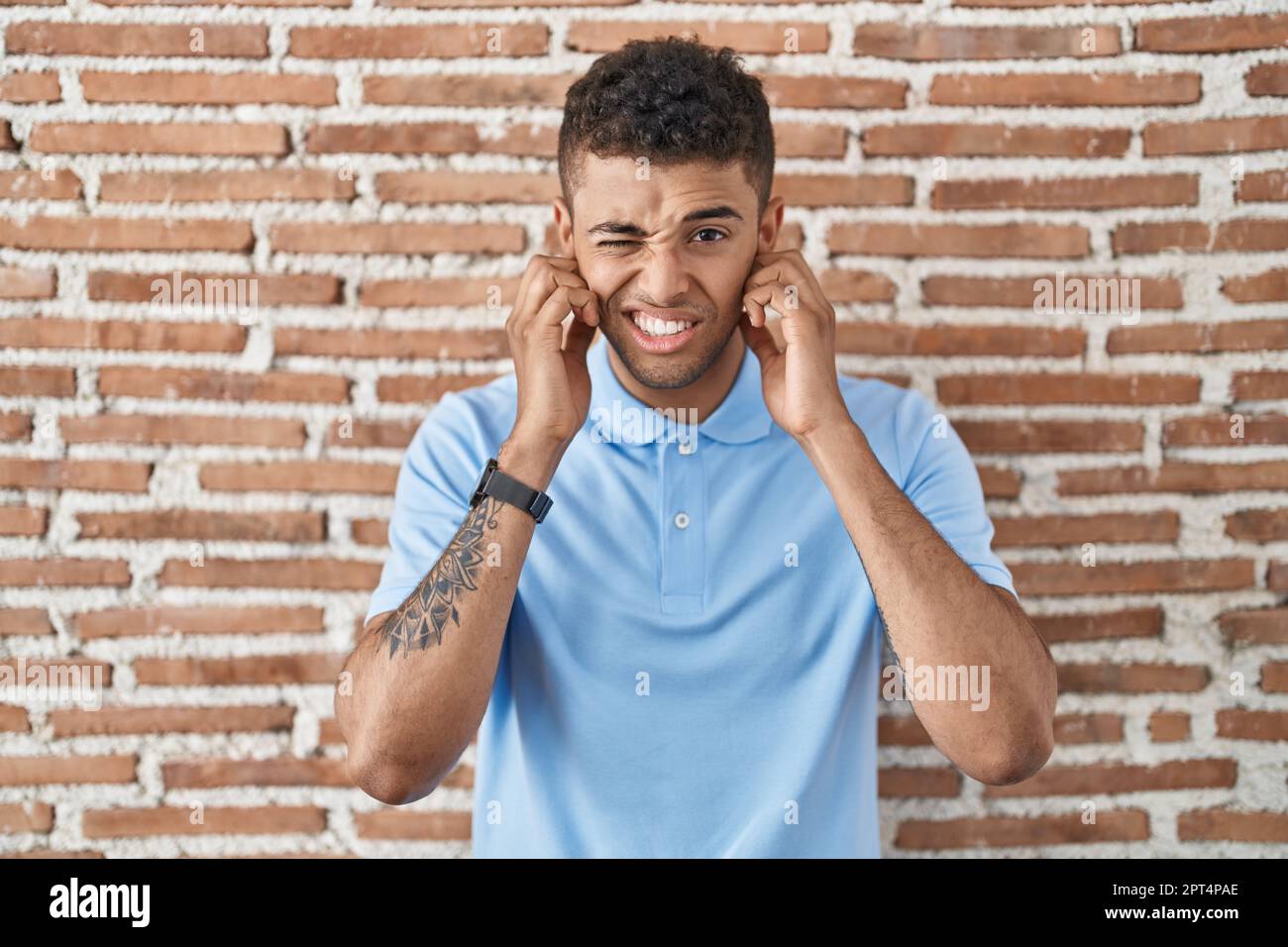 Brazilian young man standing over brick wall covering ears with fingers ...