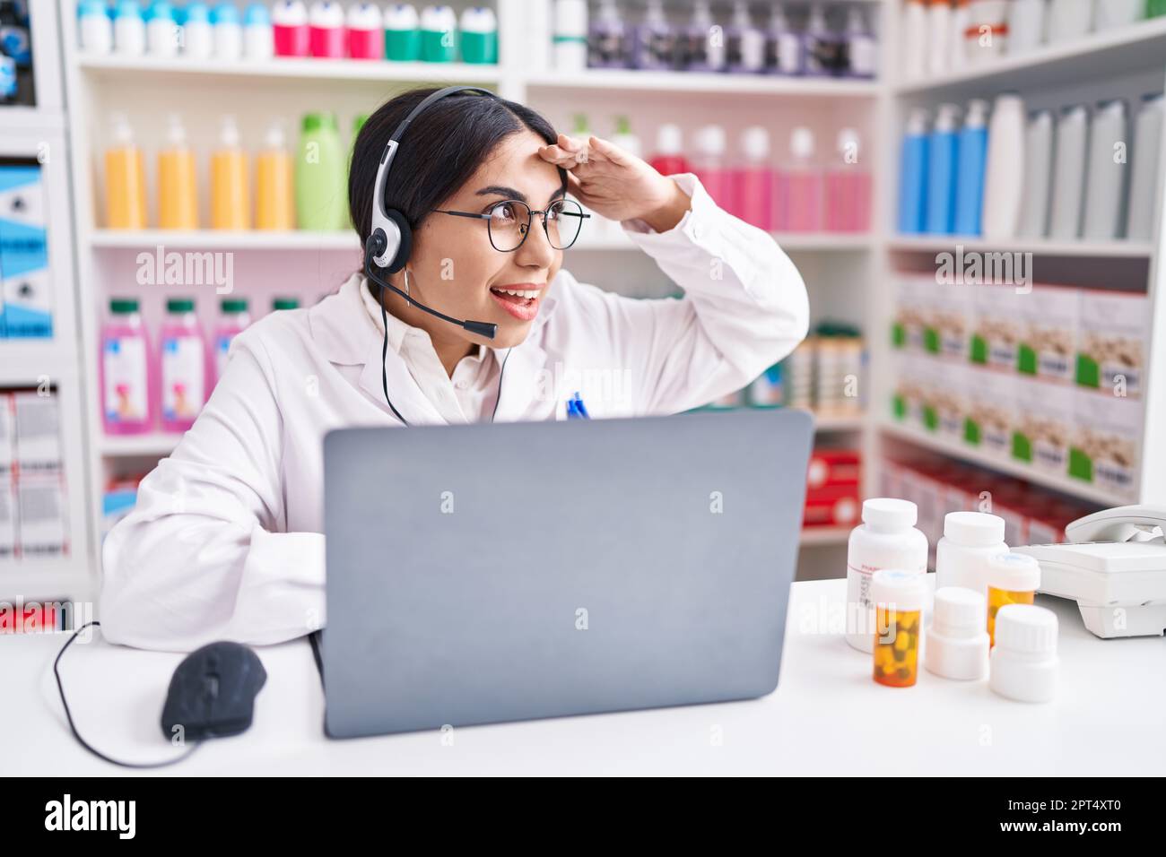 Young arab woman working at pharmacy drugstore using laptop very happy and smiling looking far ...