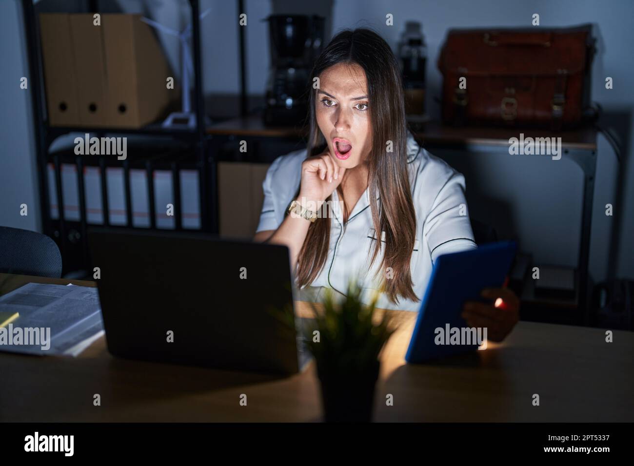 Young brunette woman working at the office at night in shock face ...