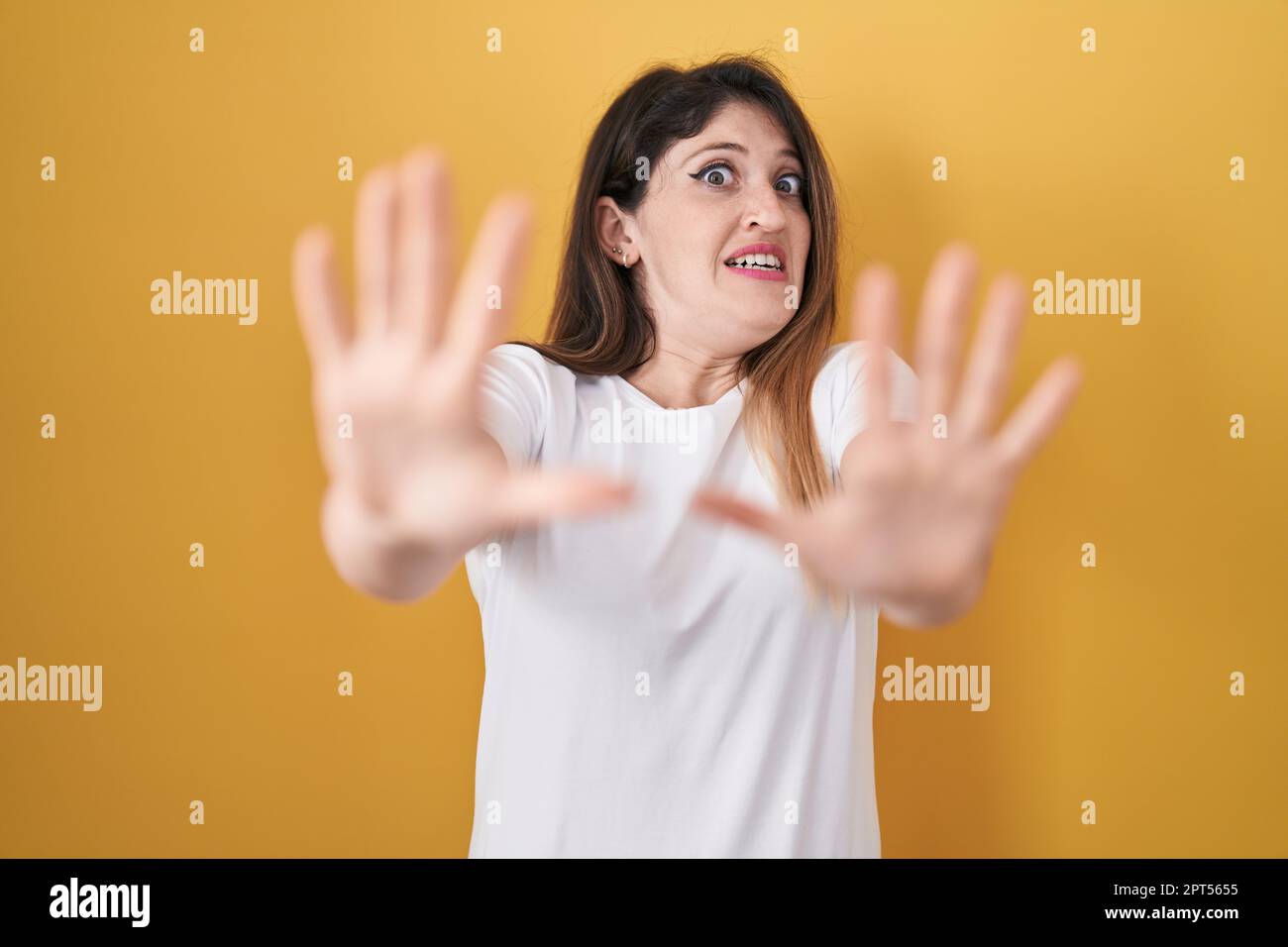 Young brunette woman standing over yellow background afraid and ...