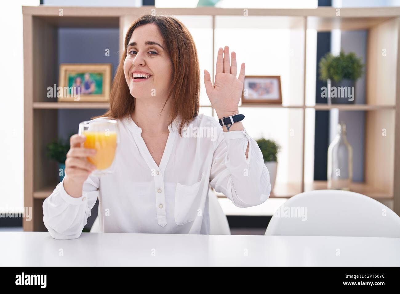 Brunette woman drinking glass of orange juice waiving saying hello ...