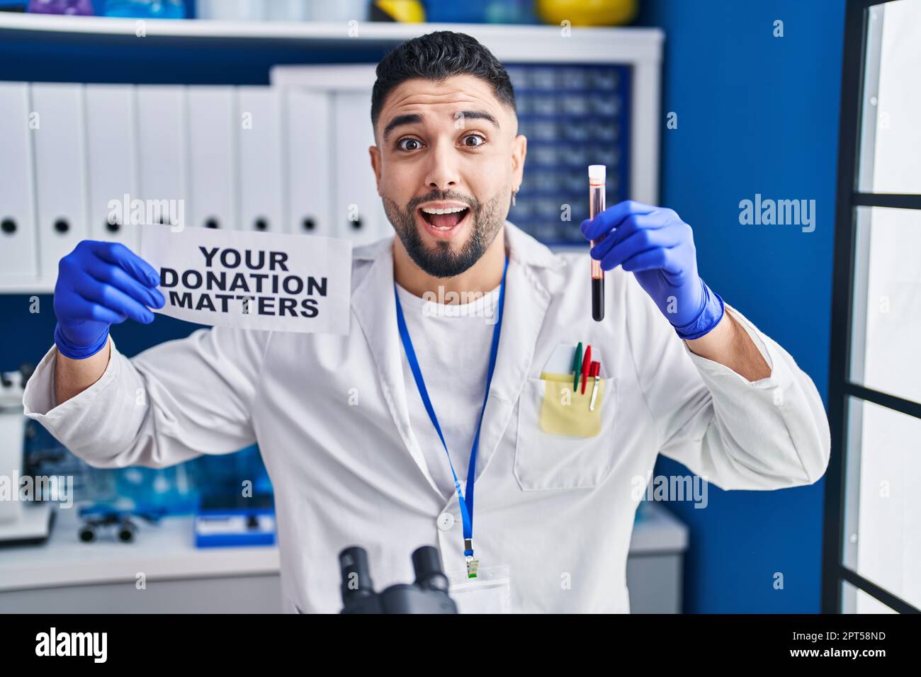 Young handsome man working at scientist laboratory holding blood sample celebrating crazy and ...