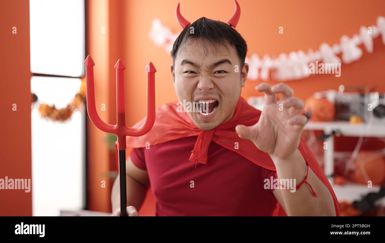 Young chinese man wearing devil costume doing scare gesture at home ...