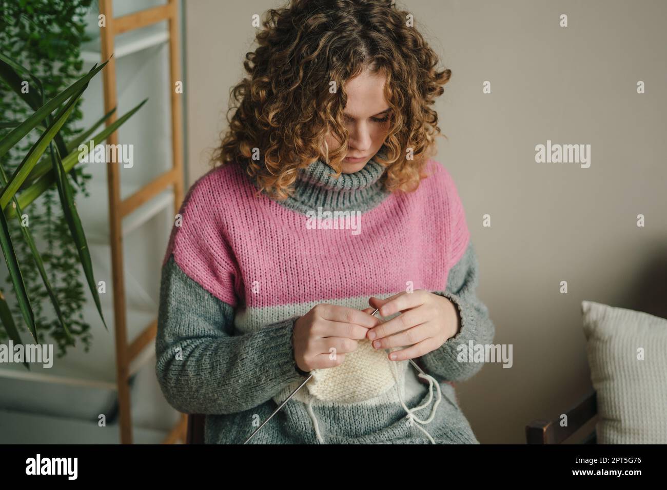 Calm curly-haired woman knitting with white thread sitting on sofa in ...
