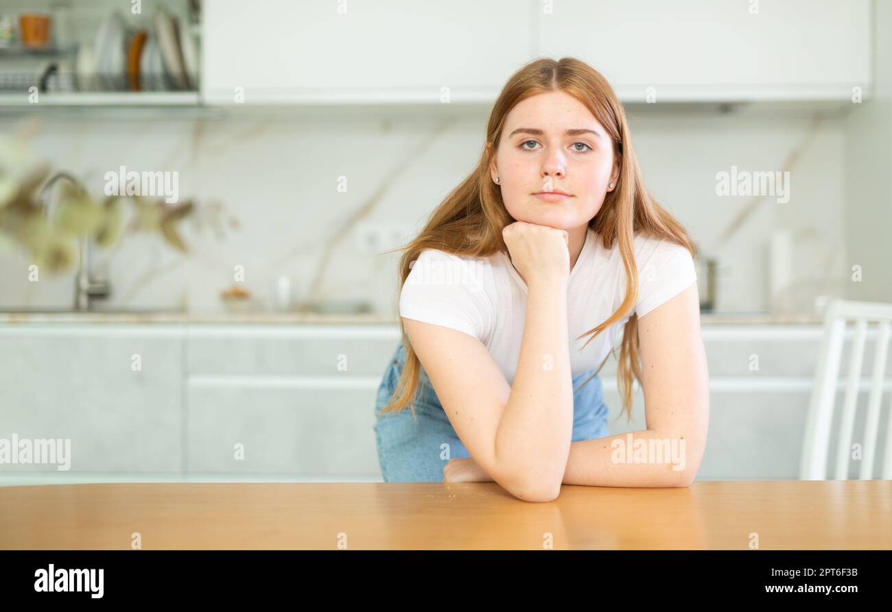 In kitchen with light furniture, long-haired girl put elbows on table ...