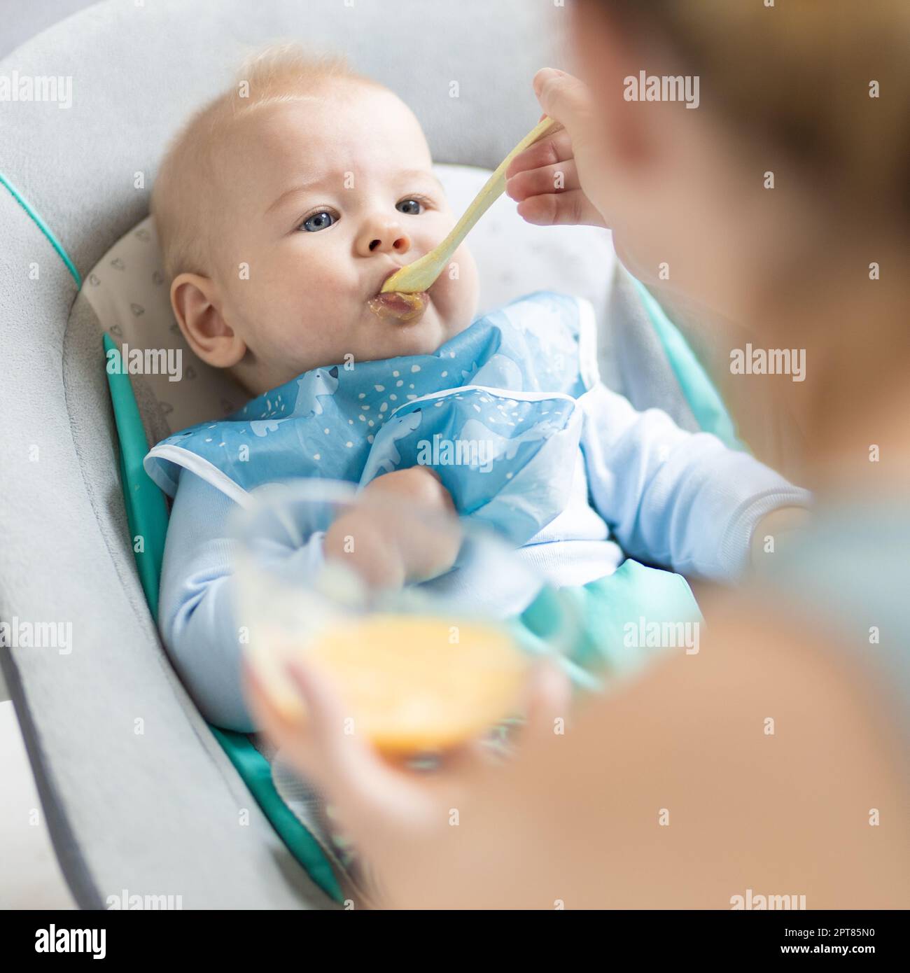 Mother spoon feeding her baby boy child in baby chair with fruit puree ...