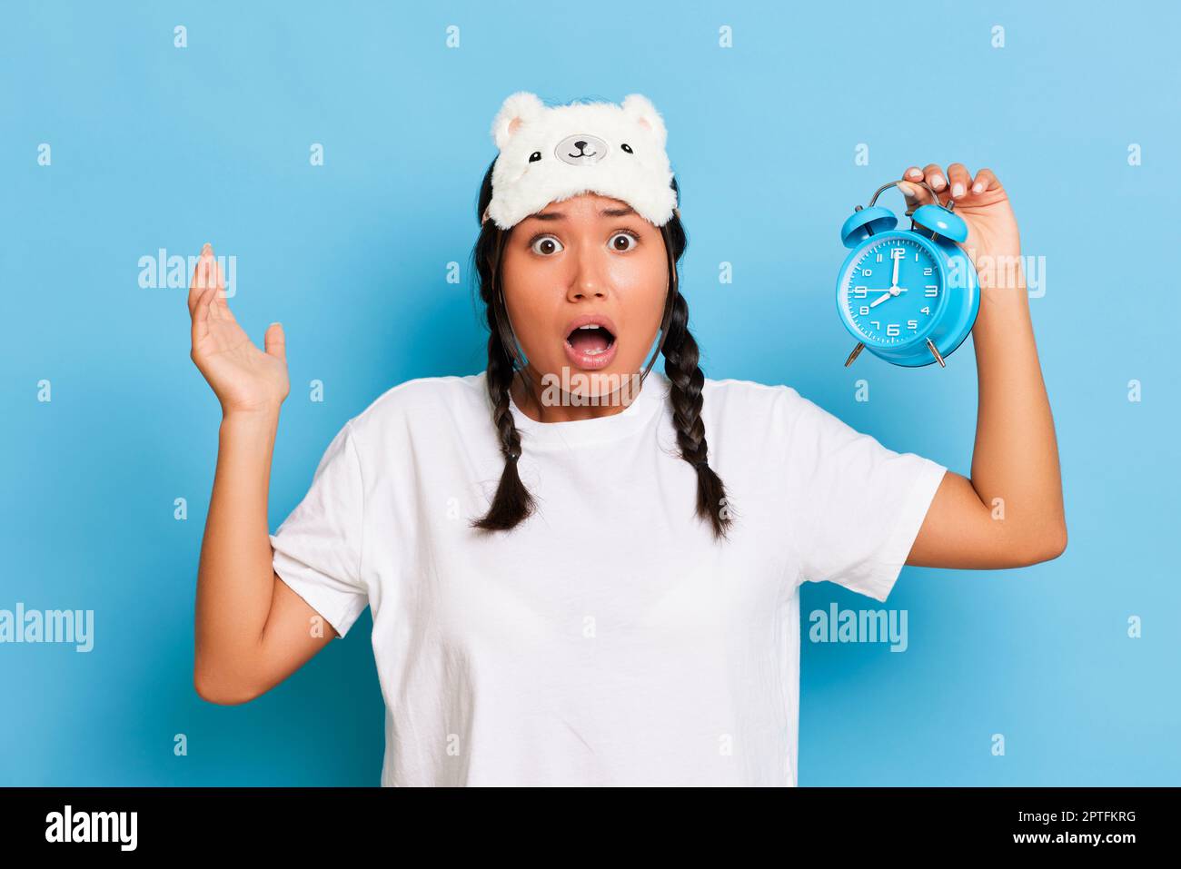 Young brunette woman with hair in plaits holding alarm clock, wears a ...