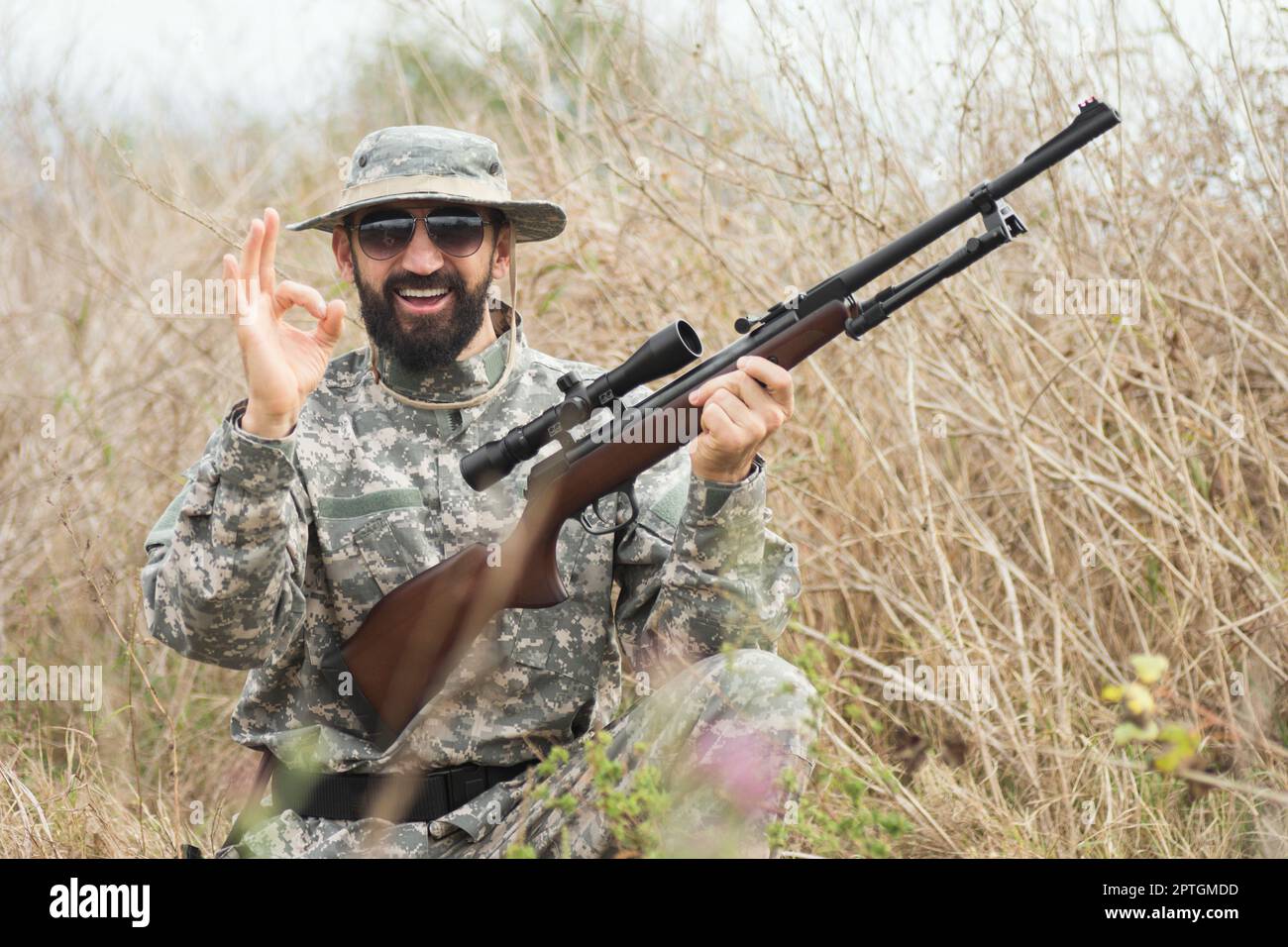 The smiling hunter in military uniform with rifle is showing OK with ...