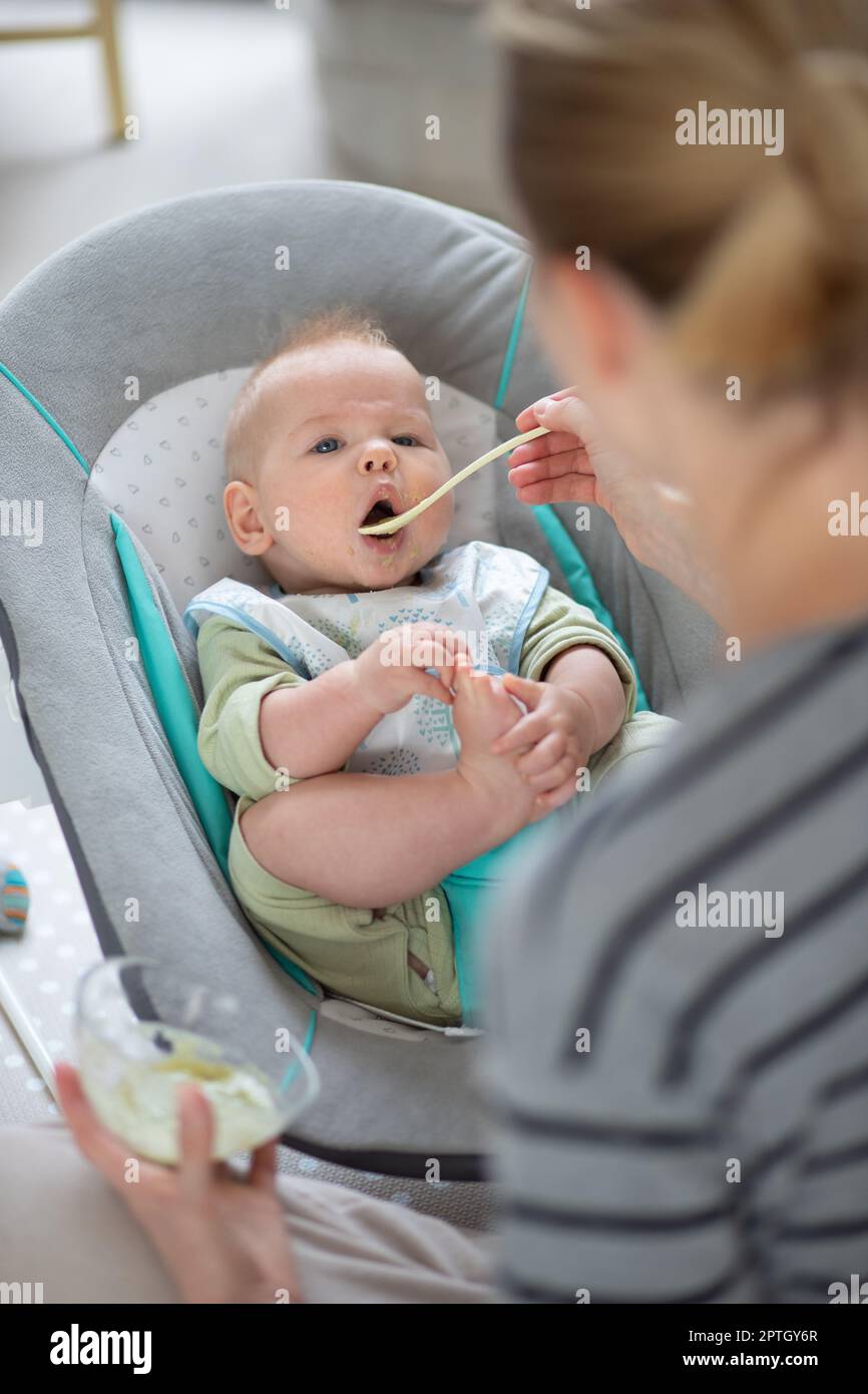Mother spoon feeding her baby boy child in baby chair with fruit puree ...