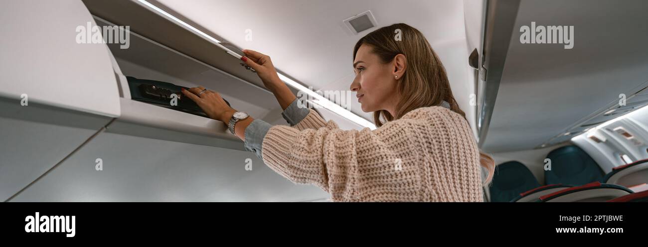 Woman traveler putting luggage into overhead locker on airplane during ...