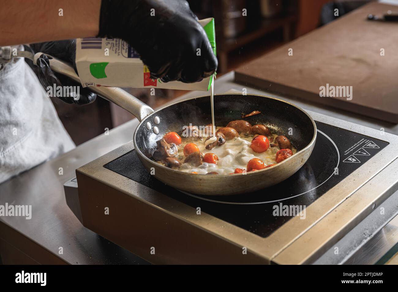 The professional chef in uniform adding cream into frying pan with dish ...
