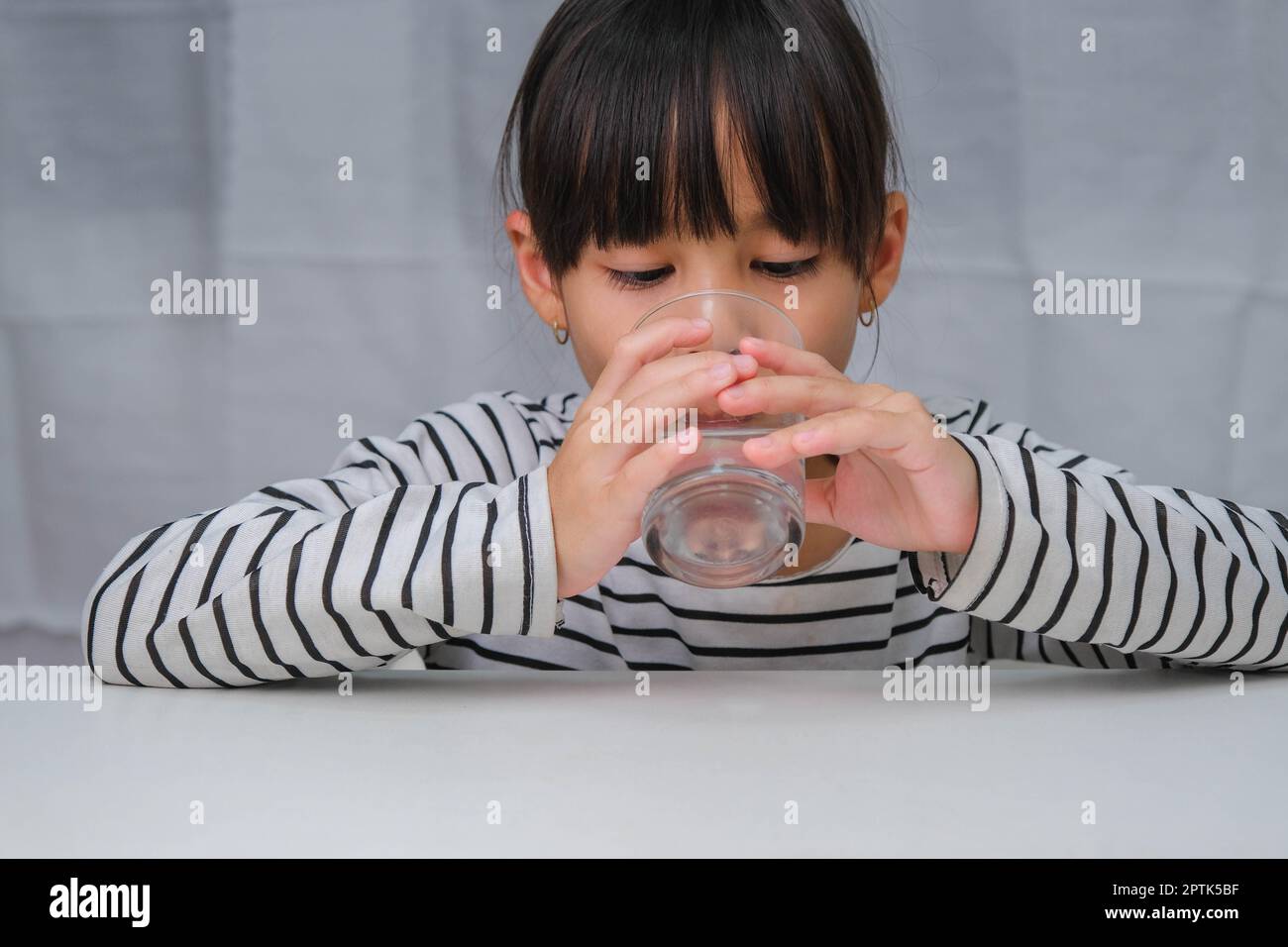 Cute little girl drinking water from glass on white curtain background ...
