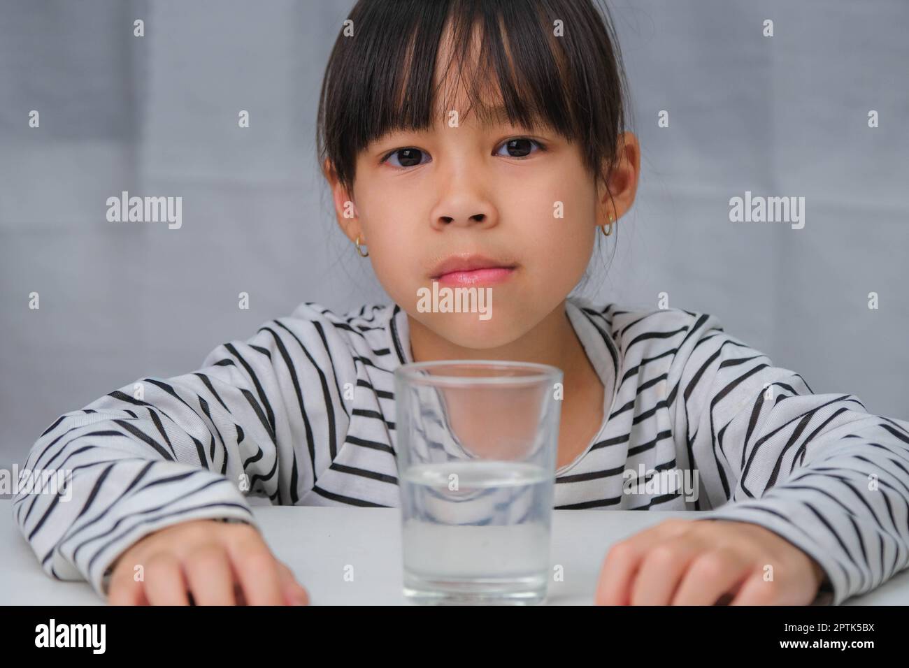 Cute little girl drinking water from glass on white curtain background. Healthy lifestyle and ...