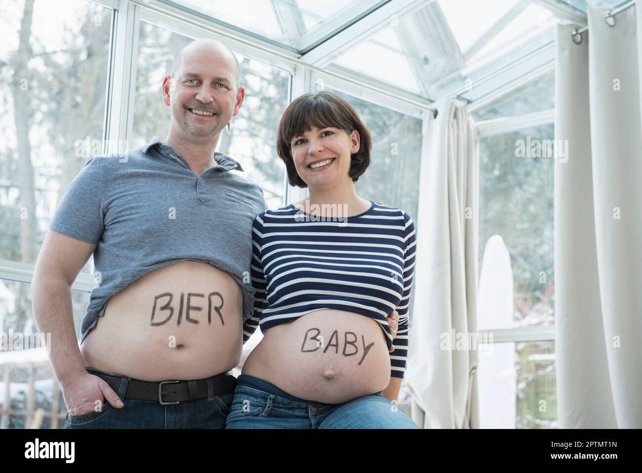 Portrait of couple showing bellies against window Stock Photo - Alamy