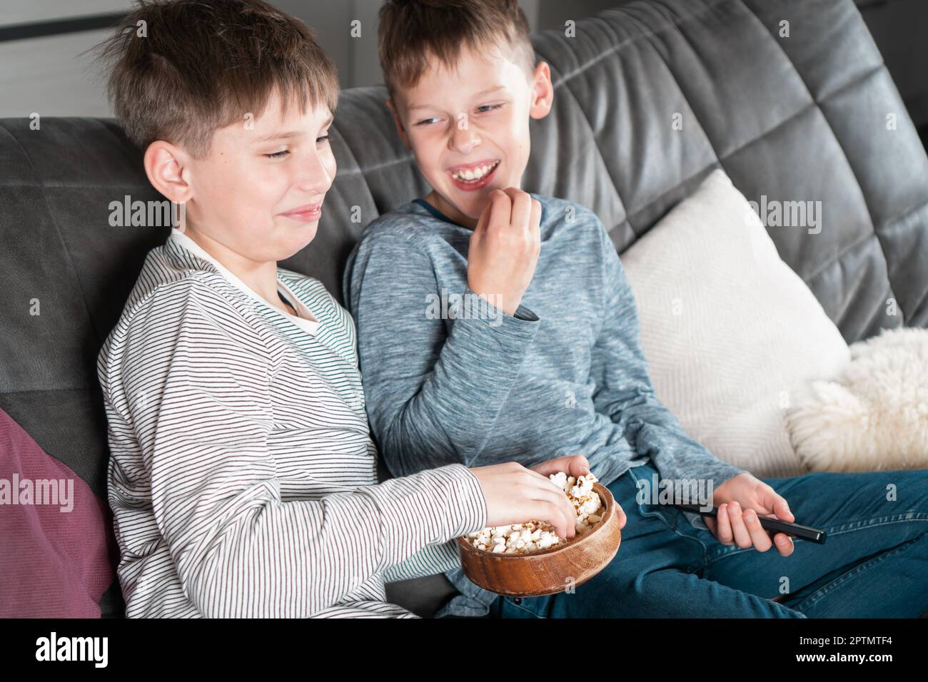 Laughing school aged boys sit on sofa, eat popcorn and watch comic TV ...