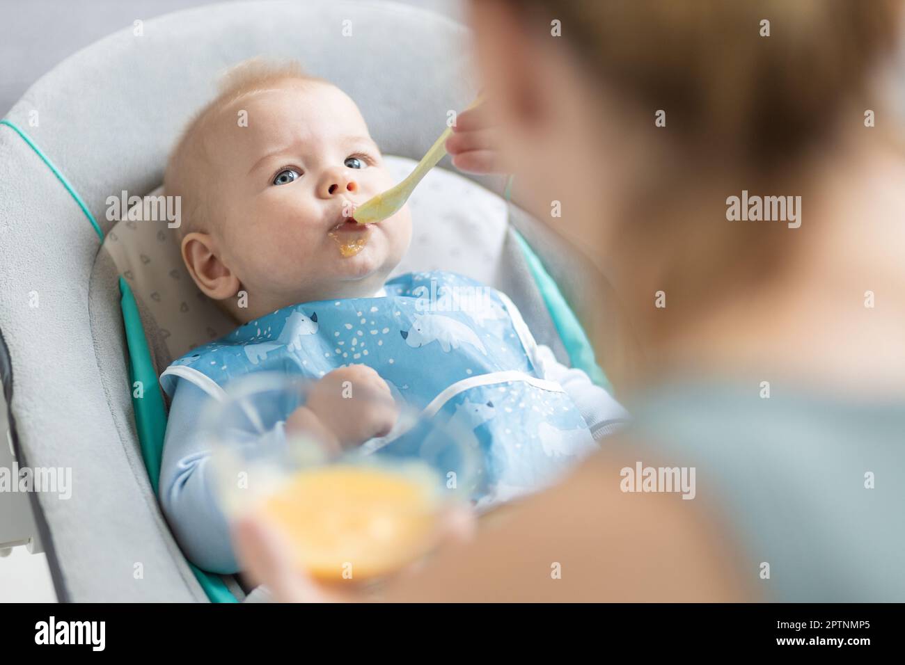 Mother spoon feeding her baby boy child in baby chair with fruit puree ...