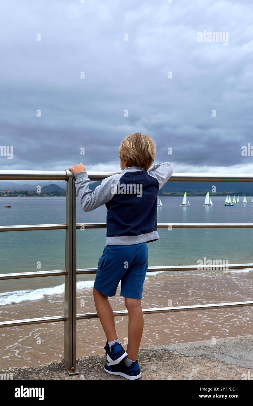 Child on his back leaning on a railing looking at sailboats. Caucasian ...