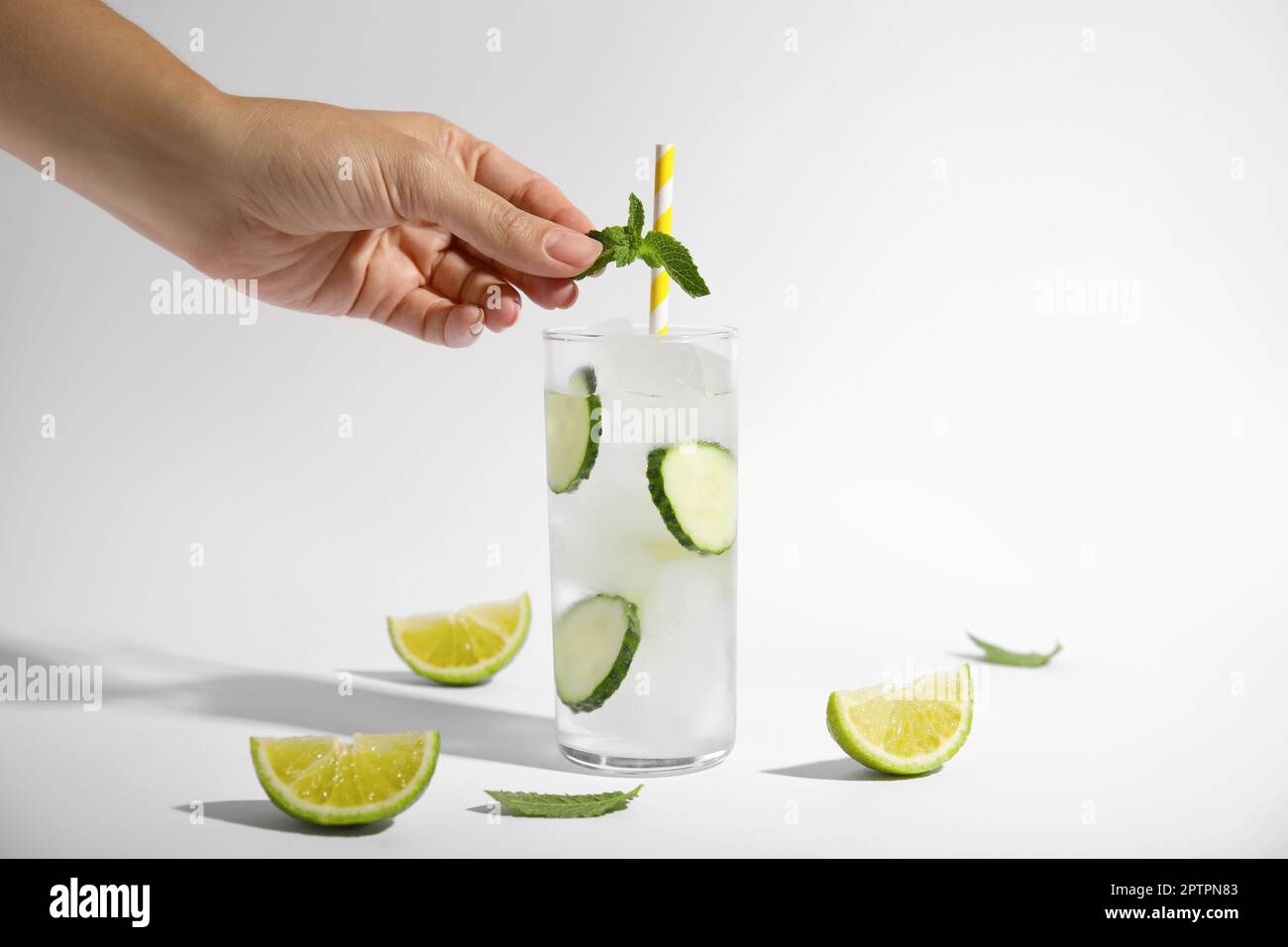 Woman putting mint into glass of tasty fresh cucumber water and sliced lime on white background ...