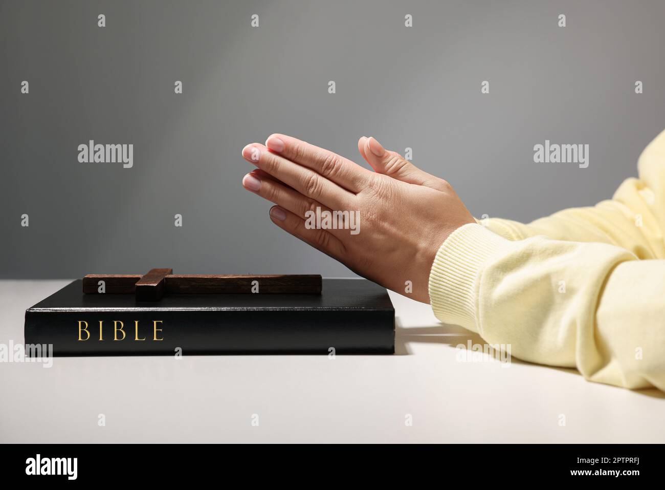 Woman praying over Bible with wooden cross at white table against grey ...