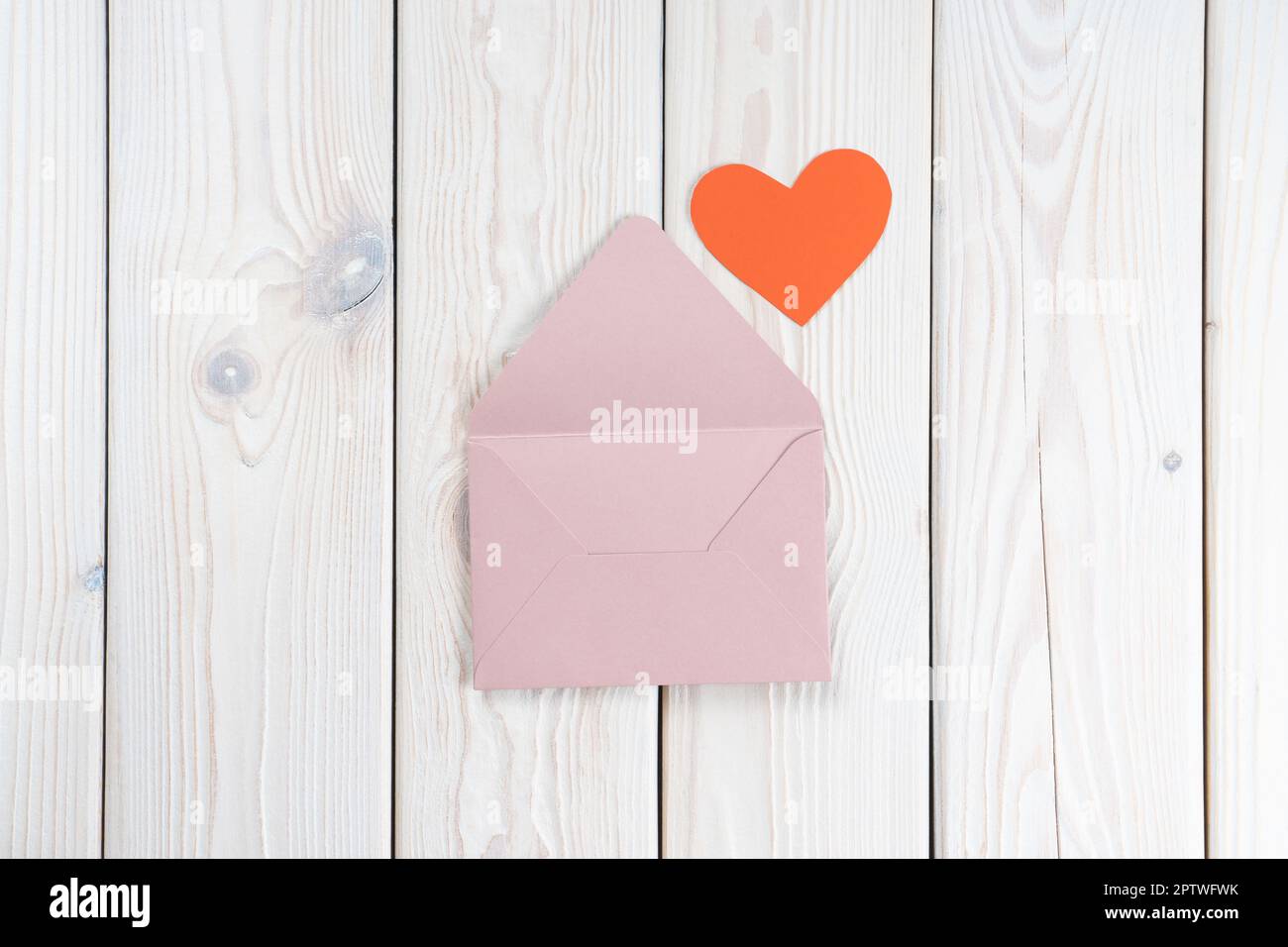 Pink opened envelope on white wooden background. Red cardboard heart is ...