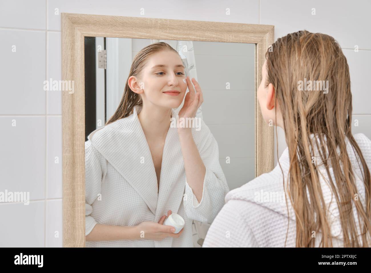 Young woman standing near the mirror and applying moisturizing cream on ...