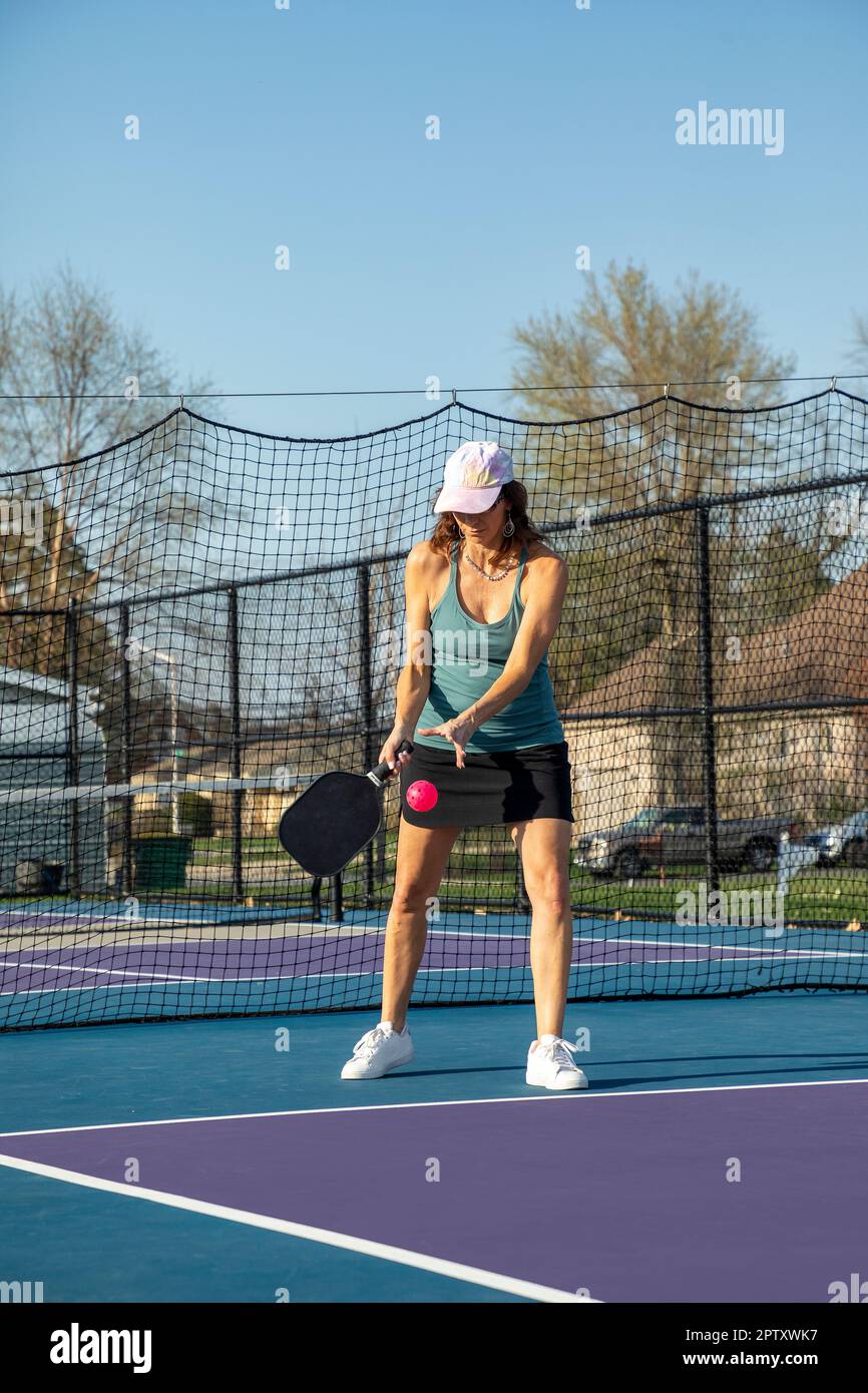 A female pickleball player serves a bright pink ball at the baseline on ...