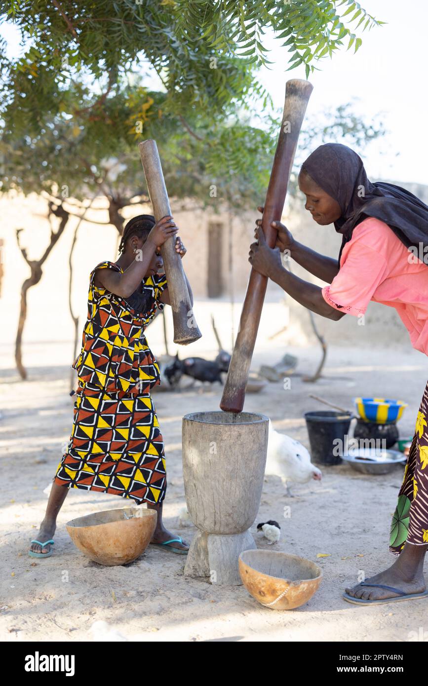A young girl and her mother pound millet grain into flour together with ...