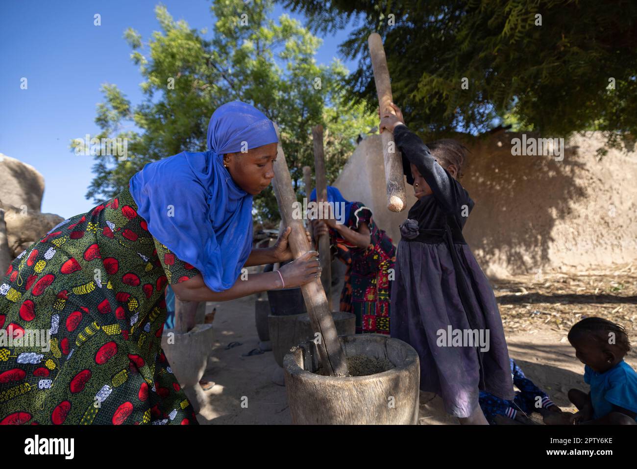 A young girl and her mother pound millet grain into flour together with ...