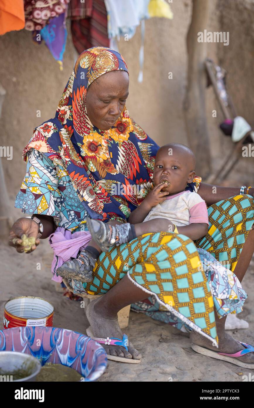 An elderly woman feeds herself and her grandchild outside her mud ...