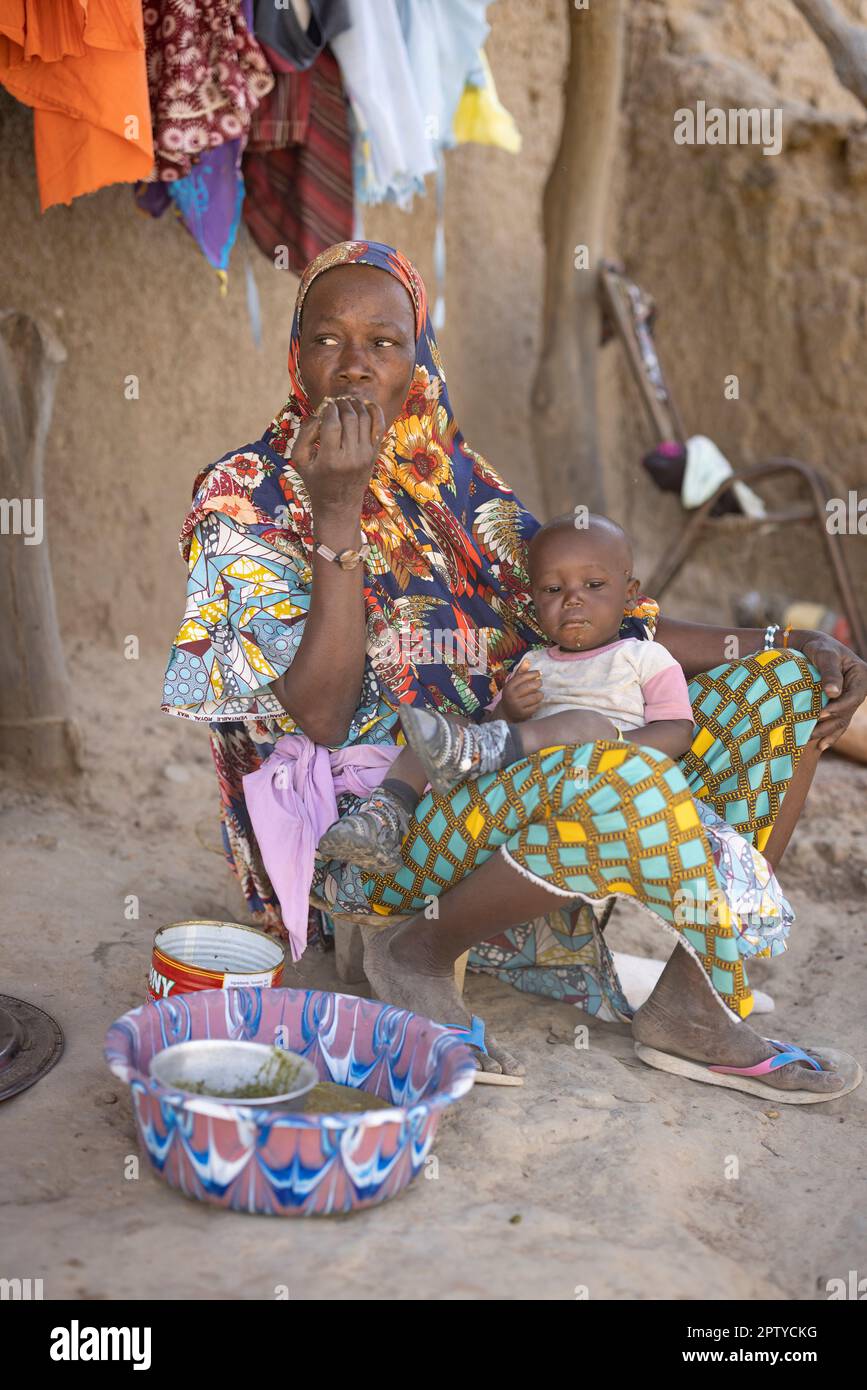 An elderly woman feeds herself and her grandchild outside her mud ...