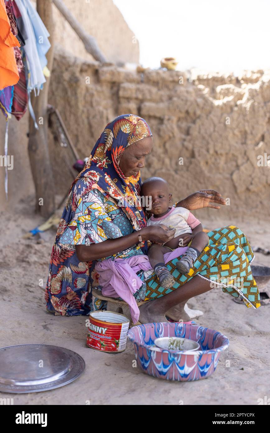 An elderly woman feeds herself and her grandchild outside her mud ...